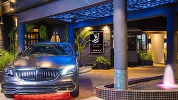 Carro de luxo estacionado em frente à entrada de um hotel, com placa, fonte de água e caminho de azulejos.