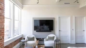 Uma sala de estar moderna com televisão de tela plana na parede, dois poltronas brancas, uma mesa de centro preta e uma parede de tijolos.