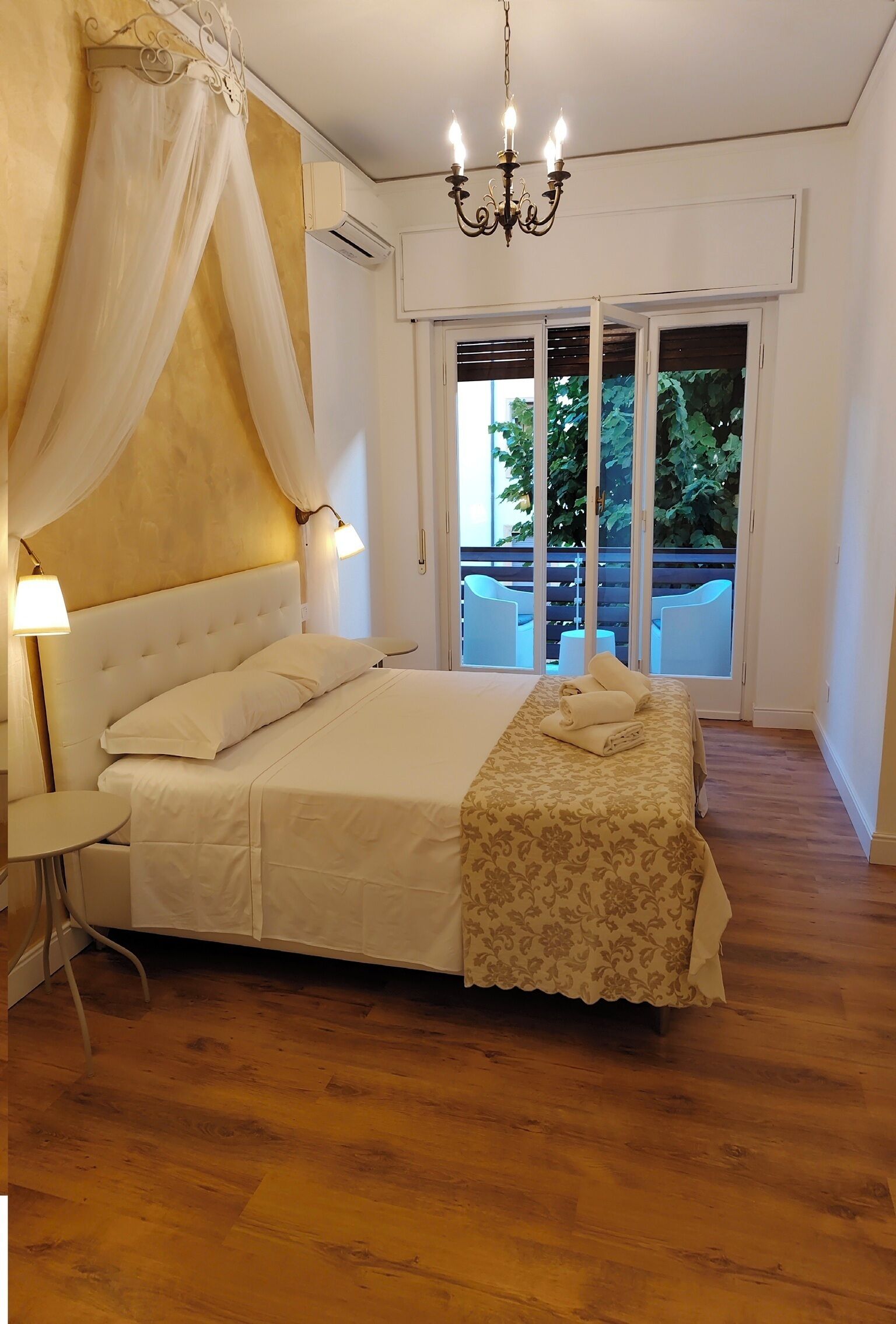 Quarto com cama grande, lustre, mesa de cabeceira e vista para o exterior através de uma porta de correr de vidro.