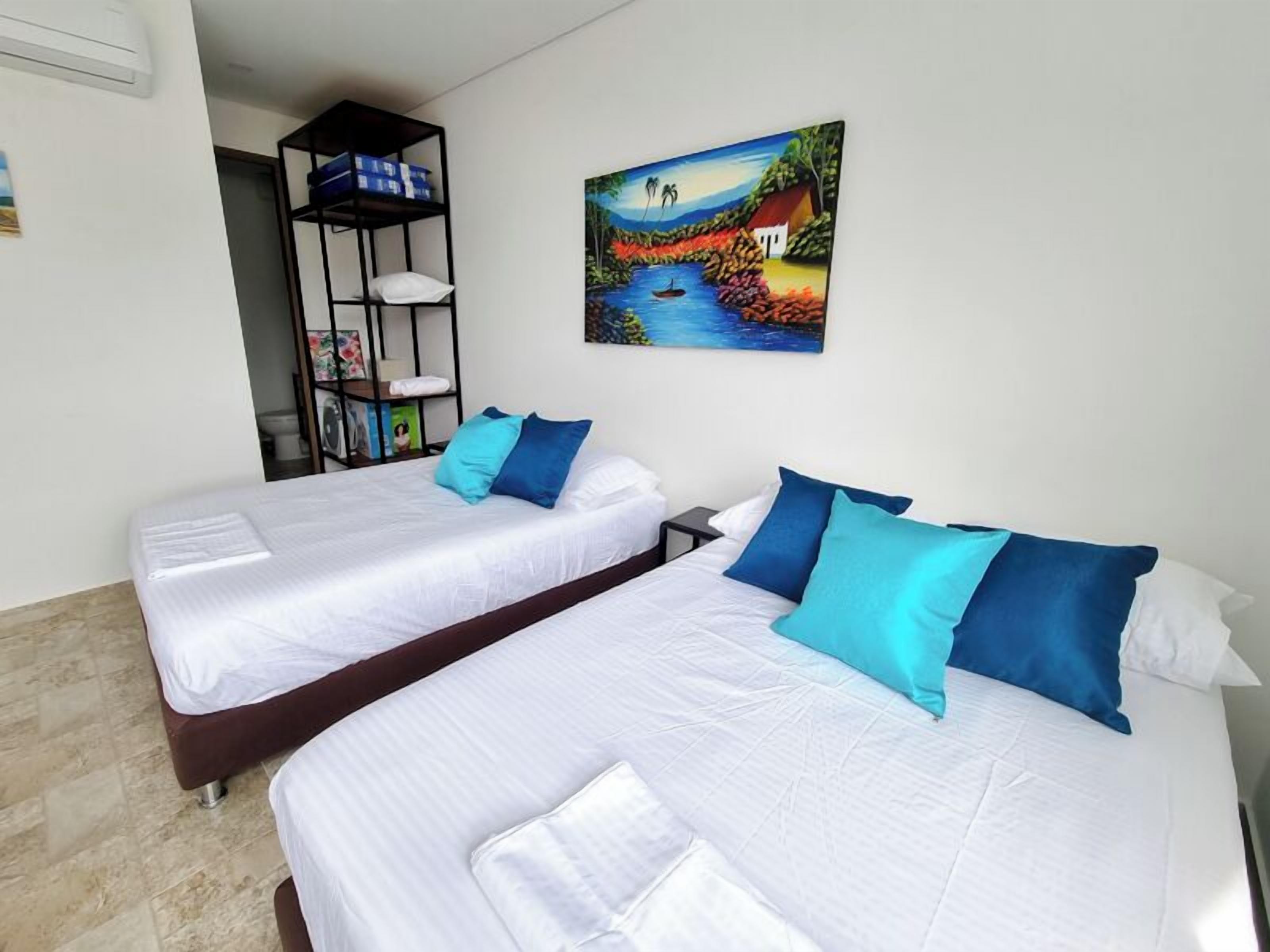 Duas camas de solteiro com roupa de cama branca e almofadas azuis, um quadro de uma paisagem tropical e uma prateleira preta com diversos objetos.