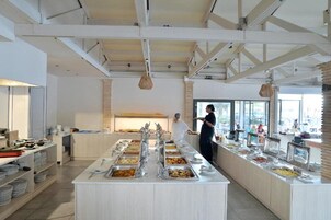 Buffet montado com diversas estações de comida, uma pessoa servindo e vista através de amplas janelas.
