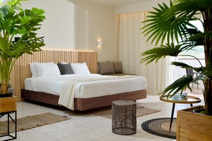 Quarto de hotel com cama, mesa de cabeceira, planta em vaso e janela com cortinas.