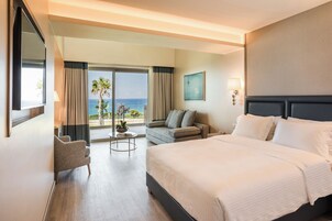Quarto de hotel com cama grande, sofá, mesa pequena com planta e vista para o oceano.