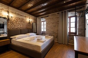 Quarto com parede de pedra, vigas de madeira no teto, cama com roupa de cama branca, mesa de cabeceira e janela com cortinas.