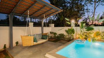Uma área de estar coberta ao ar livre com uma cadeira de balanço, piscina e vegetação ao redor.