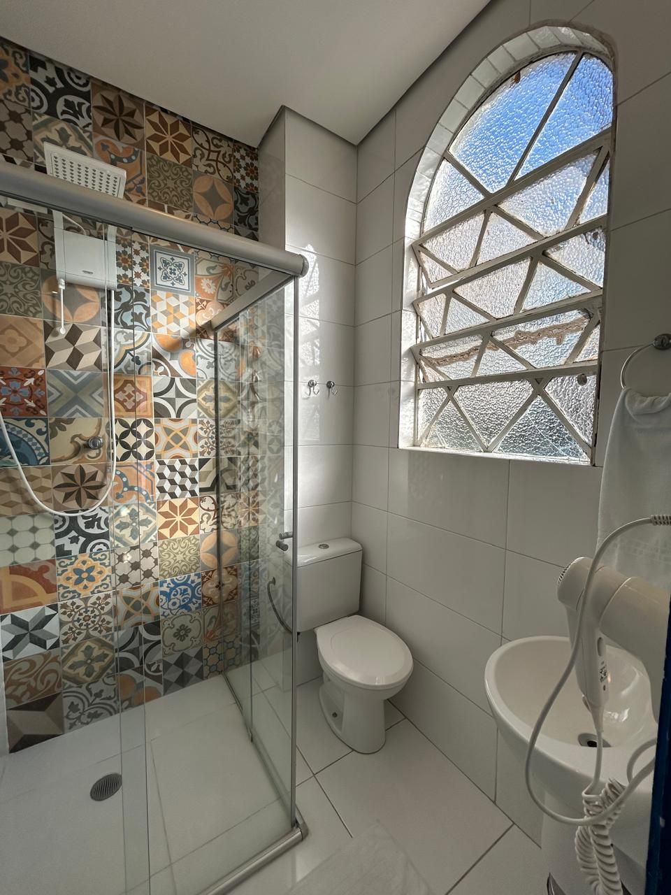 Casa de banho com chuveiro, sanita e lavatório, com azulejos estampados e uma janela grande.