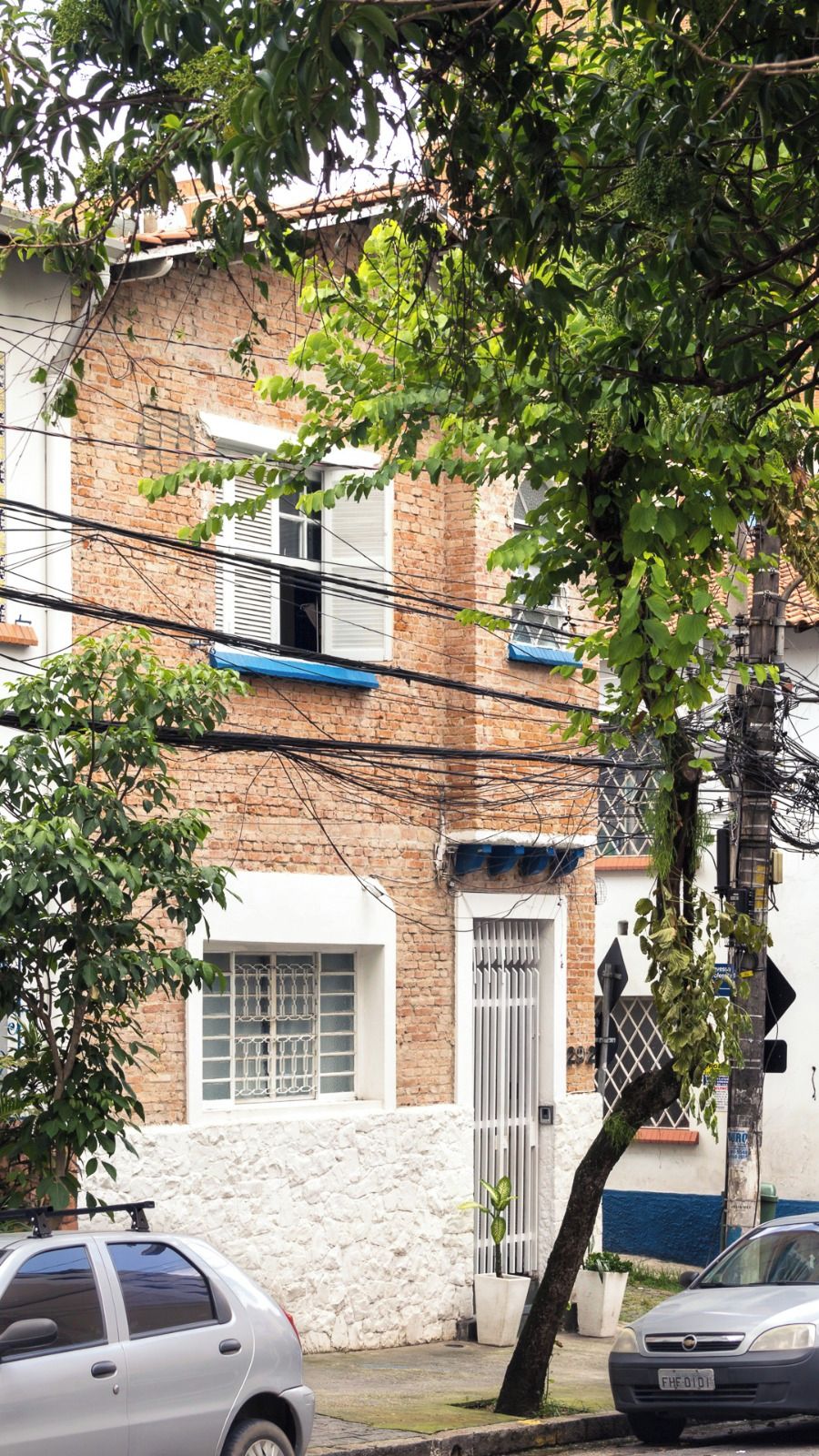 Um edifício residencial com paredes de tijolo, uma porta branca e uma janela com grade de metal. Carros estão estacionados na frente e uma árvore com folhas verdes no primeiro plano.