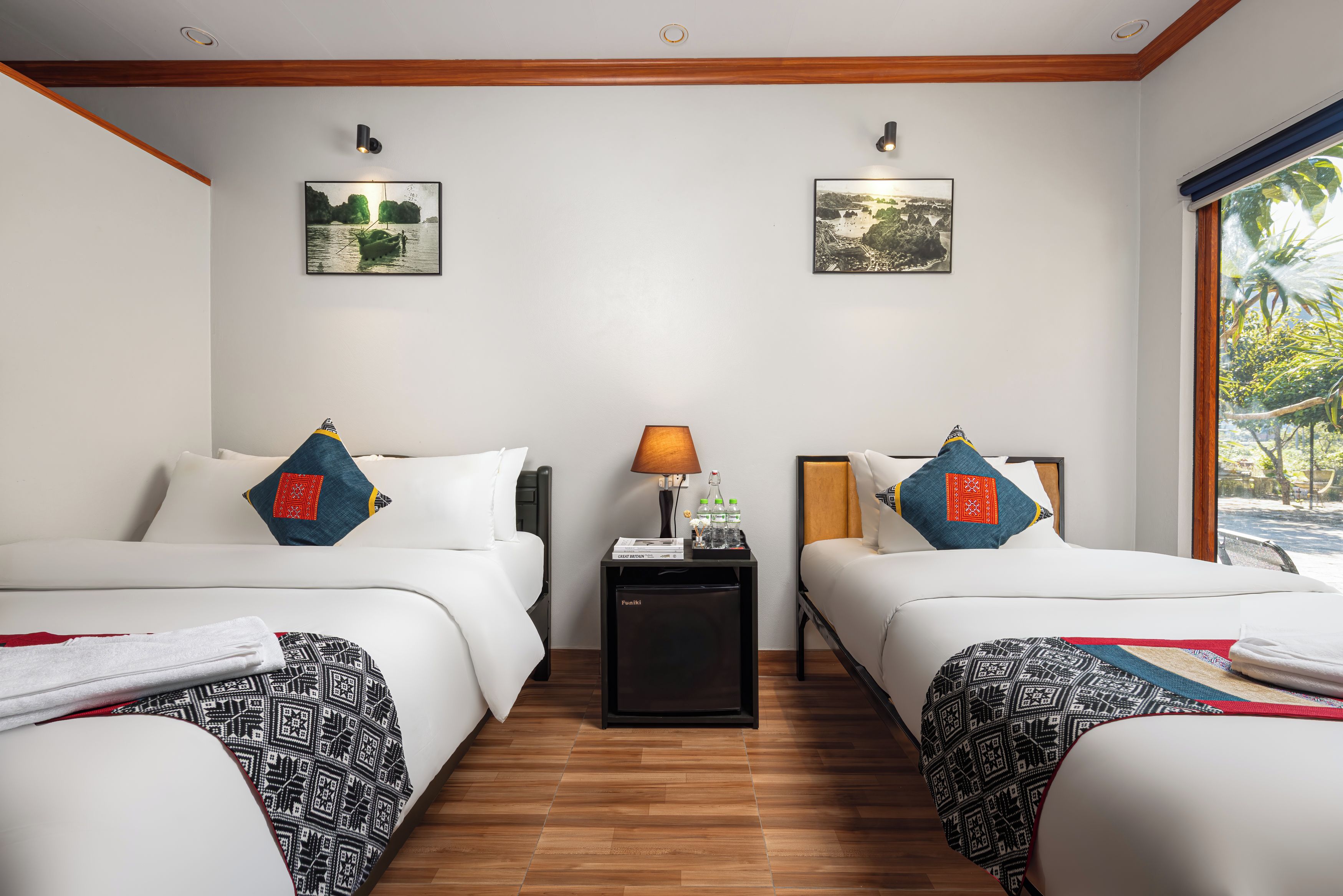 Quarto de hotel com duas camas, uma mesa de cabeceira, uma luminária e duas fotografias emolduradas na parede.