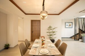 Uma sala de jantar com uma mesa comprida posta para seis pessoas, um candelabro e uma escadaria ao fundo.