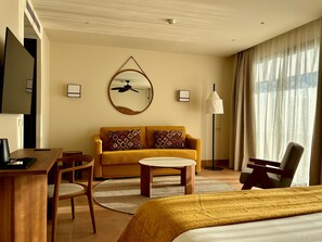 Quarto de hotel com cama, sofá, escrivaninha e uma cadeira. Há um ventilador de teto, uma luminária e uma TV fixada na parede.