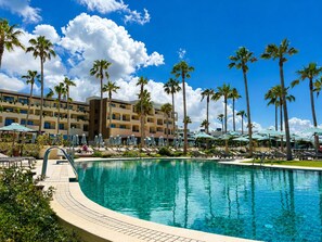 Área de piscina em resort, com palmeiras, espreguiçadeiras e um prédio com várias varandas.