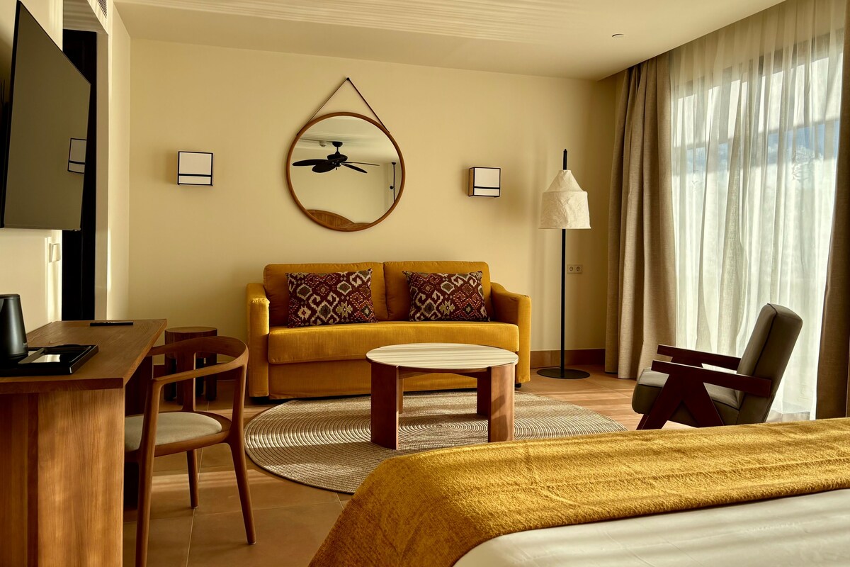 Quarto de hotel com cama, sofá, escrivaninha e uma cadeira. Há um ventilador de teto, uma luminária e uma TV fixada na parede.