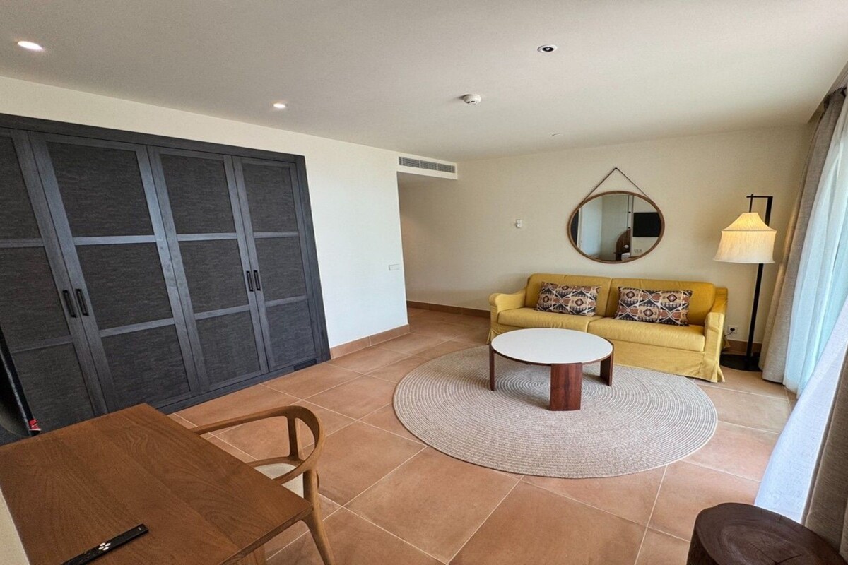 Sala de estar com sofá, mesa de centro e mesa de jantar com cadeiras. Há um guarda-roupa grande e um espelho na parede.