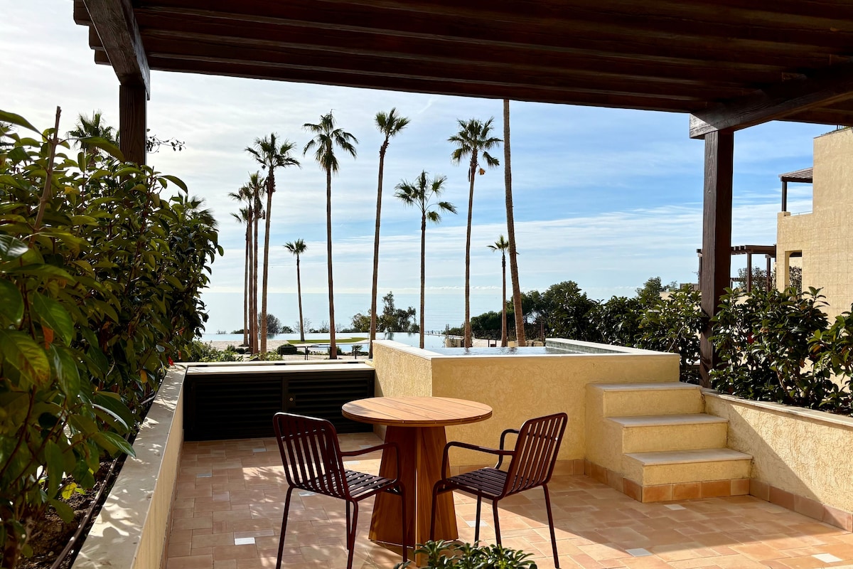 Uma área de estar coberta ao ar livre com mesa e cadeiras, com vista para uma piscina e palmeiras.