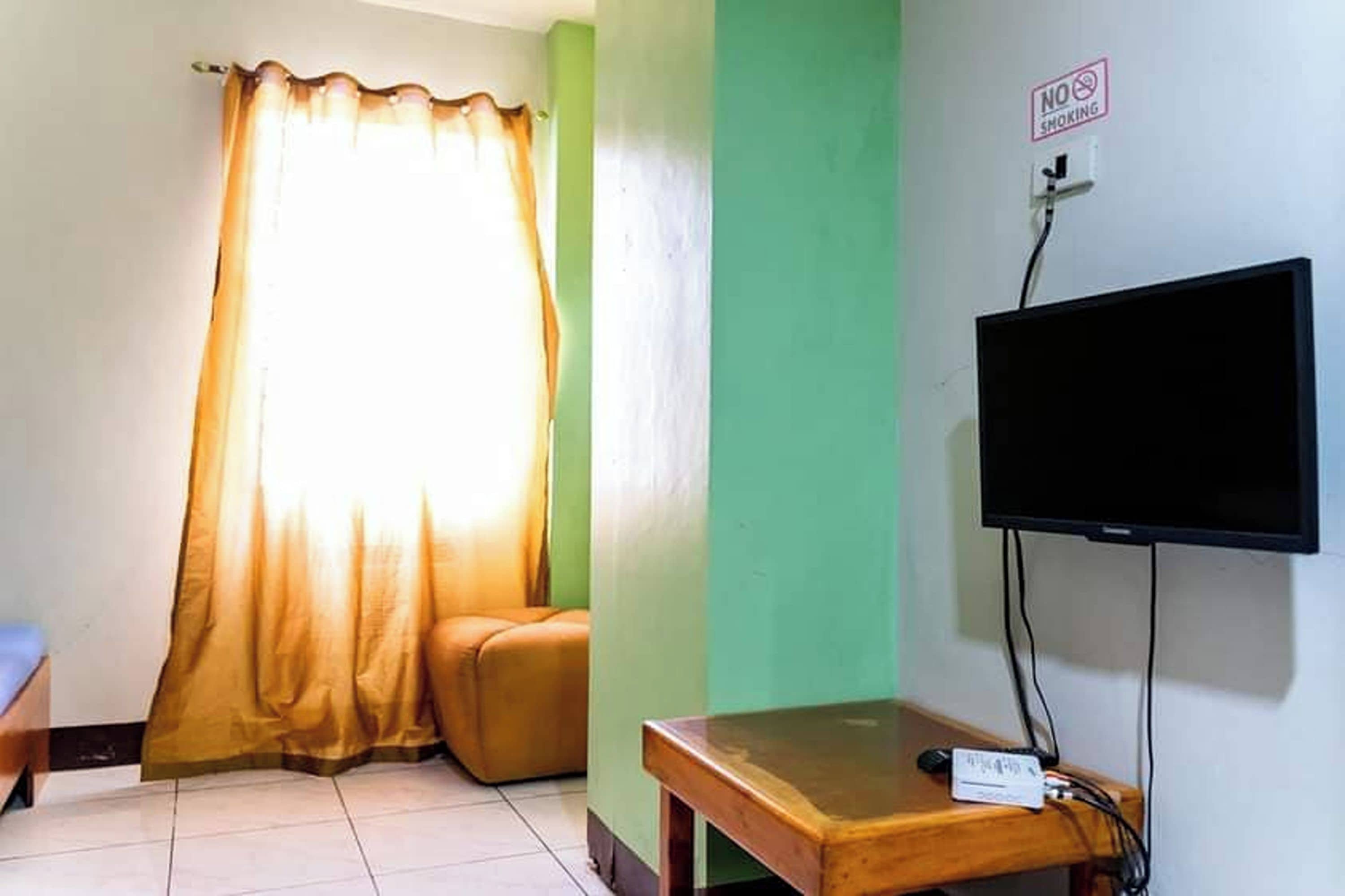 Quarto com televisão na parede, uma mesa de madeira, sofá e janela com cortinas.