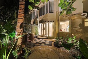 Área externa bem iluminada, com uma palmeira, um elemento de água e um caminho que leva a um edifício com grandes janelas.