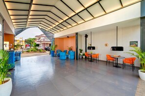 Amplo espaço ao ar livre com design moderno, incluindo área de estar coberta com cadeiras azuis e laranjas, uma mesa com tabuleiro de xadrez e plantas em vasos.