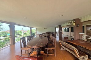 Uma sala de jantar espaçosa com uma grande mesa de madeira e cadeiras, uma varanda com guarda-corpo e vista para áreas verdes e edifícios.