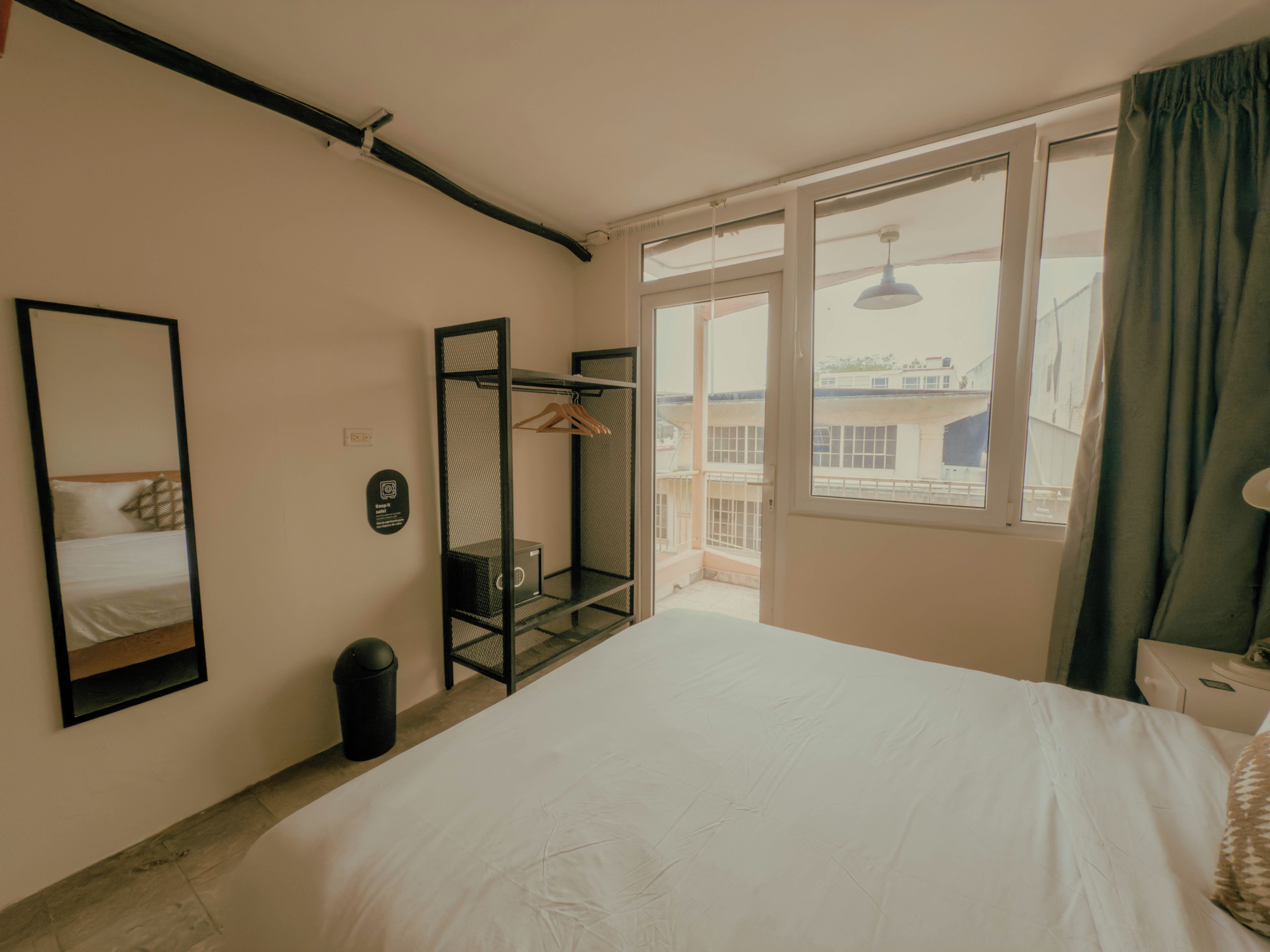 Quarto de hotel com cama, espelho, porta-casacos e janela com vista para edifícios.
