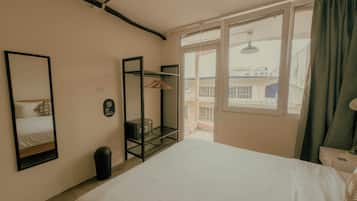Quarto de hotel com cama, espelho, porta-casacos e janela com vista para edifícios.