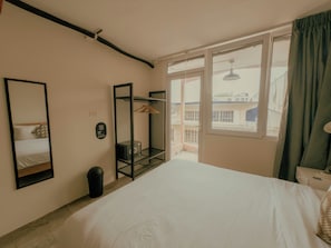 Quarto de hotel com cama, espelho, porta-casacos e janela com vista para edifícios.