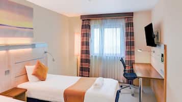 Quarto de hotel com uma cama, escrivaninha, cadeira, televisão e janela com cortinas.