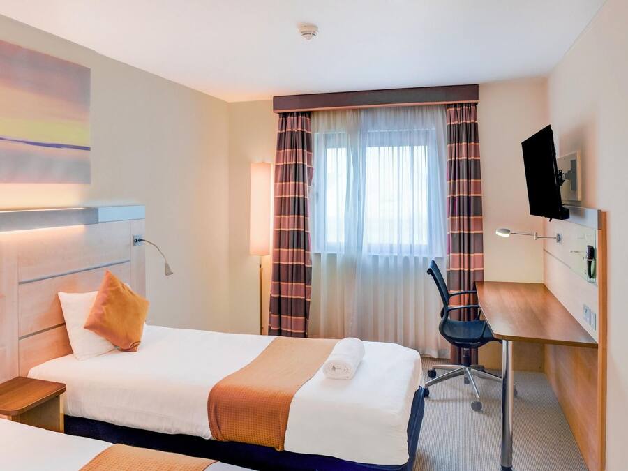 Quarto de hotel com uma cama, escrivaninha, cadeira, televisão e janela com cortinas.