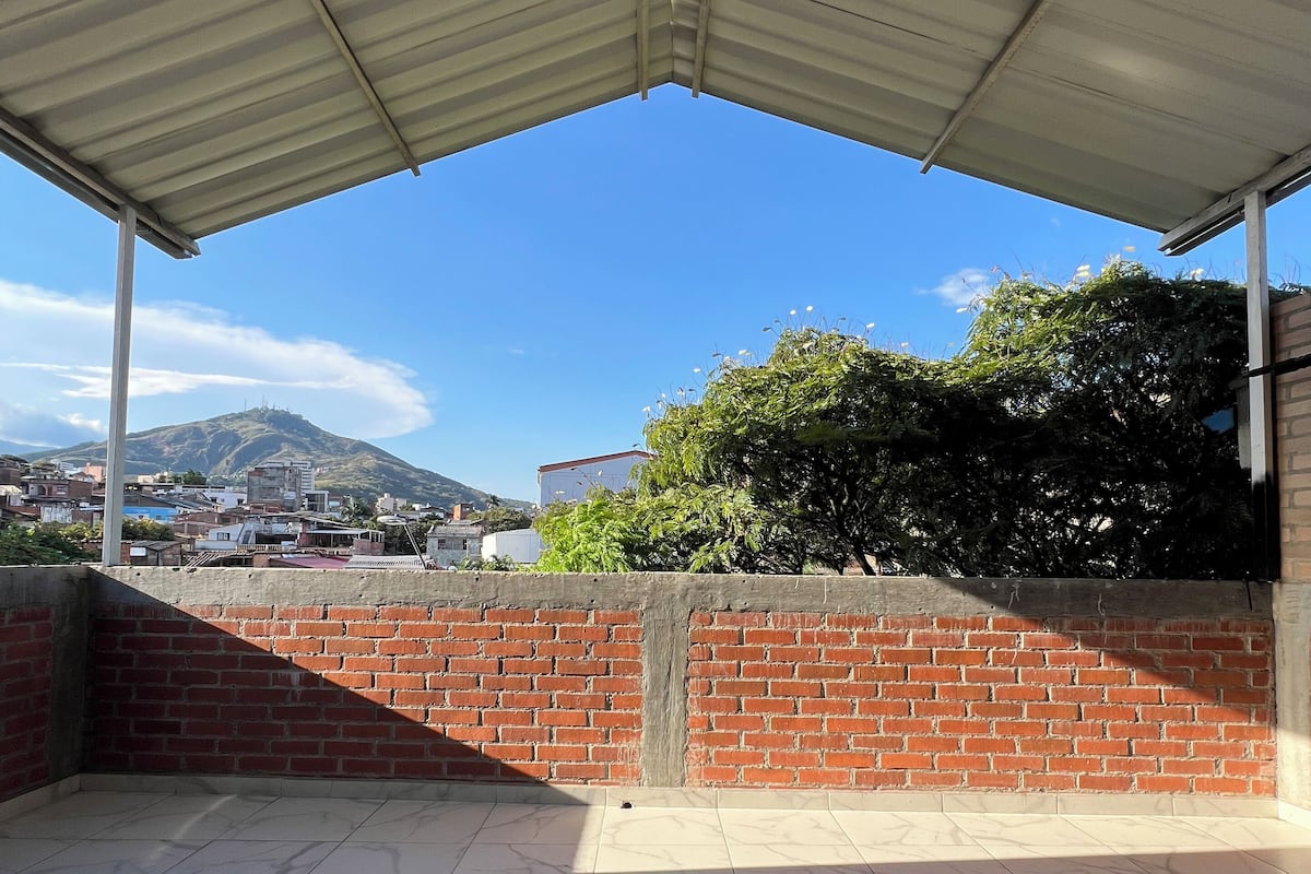 Área externa coberta com piso de azulejos, paredes de tijolo e vista para um horizonte urbano e colinas ao fundo.