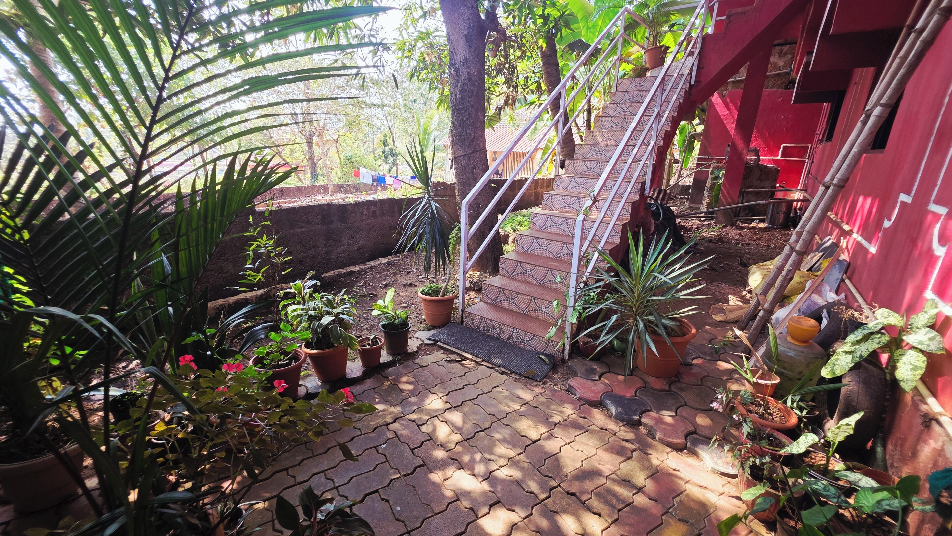 Uma trilha pavimentada conduz a uma escadaria, decorada com vasos de plantas, e um edifício vermelho ao fundo.