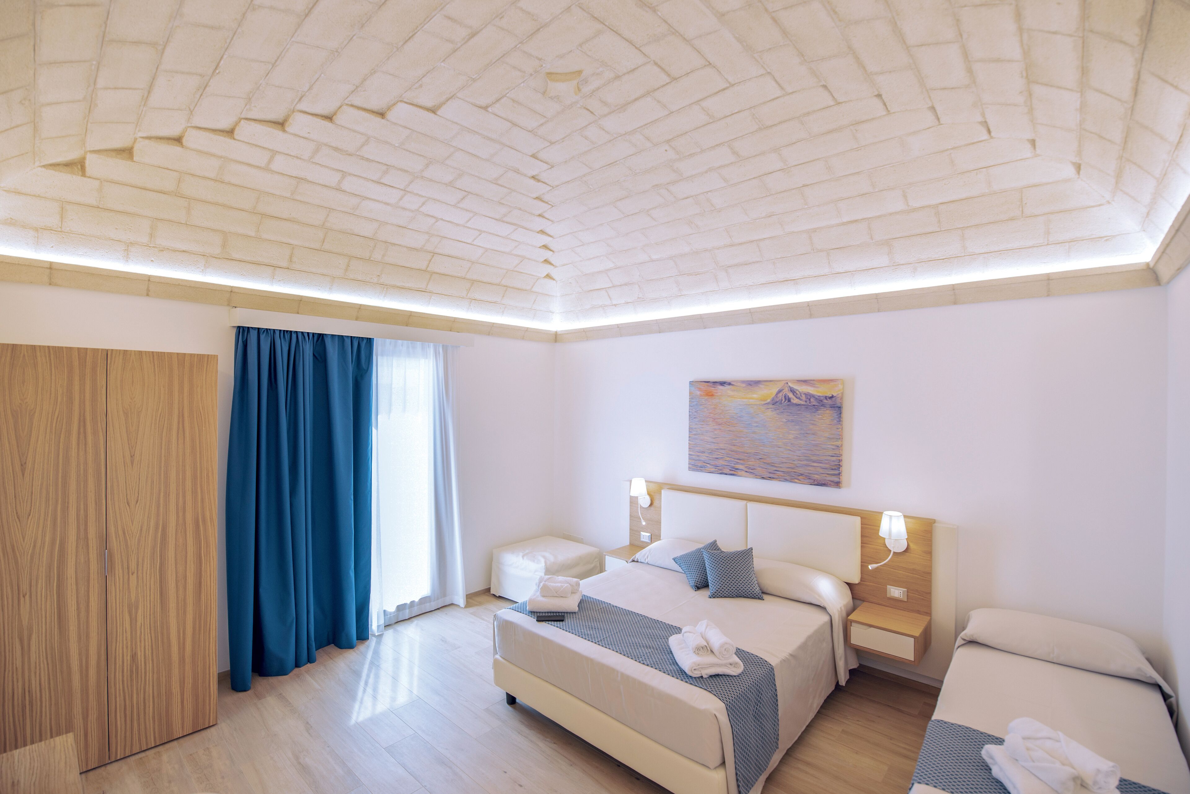 Quarto de hotel com teto em padrão de tijolos, uma cama grande com duas almofadas, uma mesa de cabeceira com abajur e um quadro na parede.