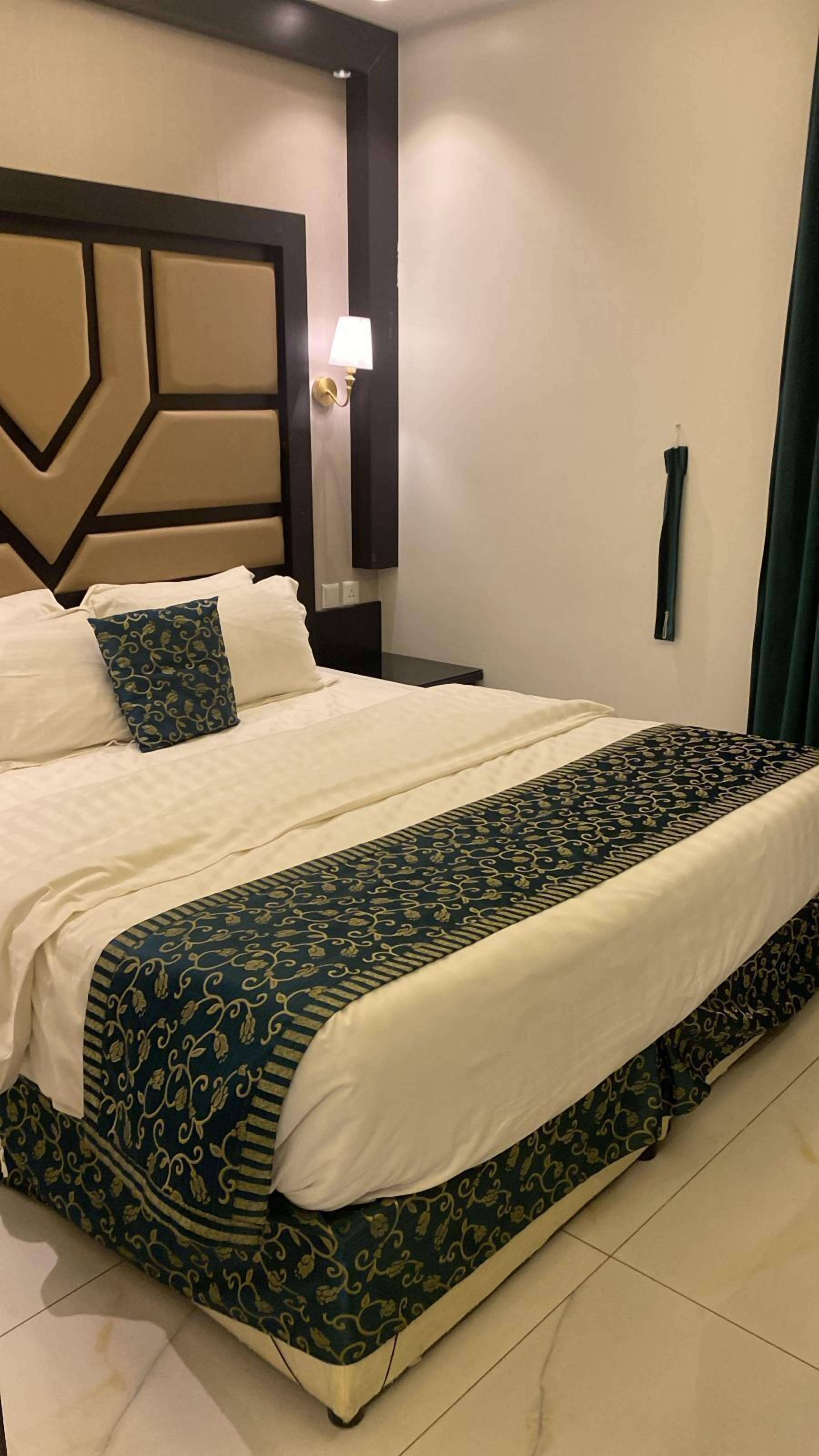 Quarto de hotel com uma cama grande, cabeceira com padrão geométrico, luminária de cabeceira e cortina escura.