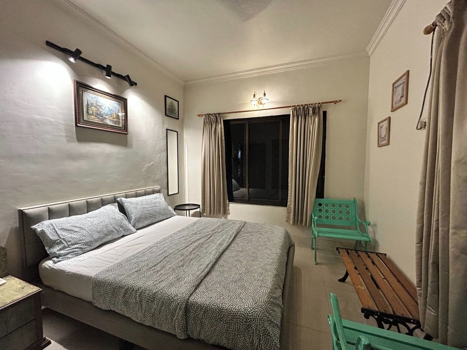 Quarto com cama, mesa de cabeceira, janela com cortinas, cadeira e um banco.