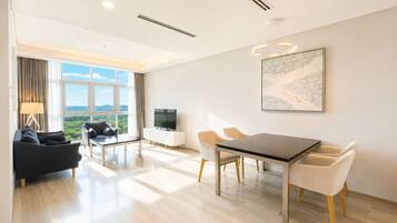 Quarto de hotel moderno com área de jantar, sofá, televisão e uma grande janela com vista panorâmica.