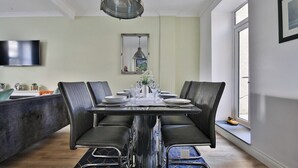 Uma área de refeições com mesa posta para quatro pessoas, rodeada por cadeiras estofadas em preto, uma luminária pendente moderna e uma grande janela que oferece luz natural.