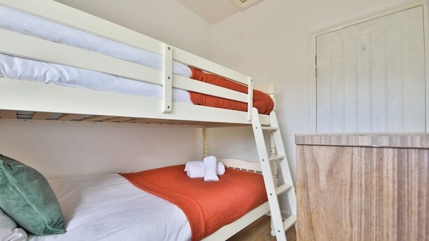 Uma cama beliche com roupa de cama branca e laranja, um travesseiro verde e um guarda-roupa de madeira ao fundo.