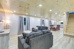 Uma sala de estar espaçosa com um sofá modular cinza grande, uma mesa de jantar com cadeiras e uma cozinha ao fundo.