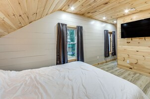 Quarto com teto de madeira, uma cama, duas janelas com cortinas e uma televisão na parede.
