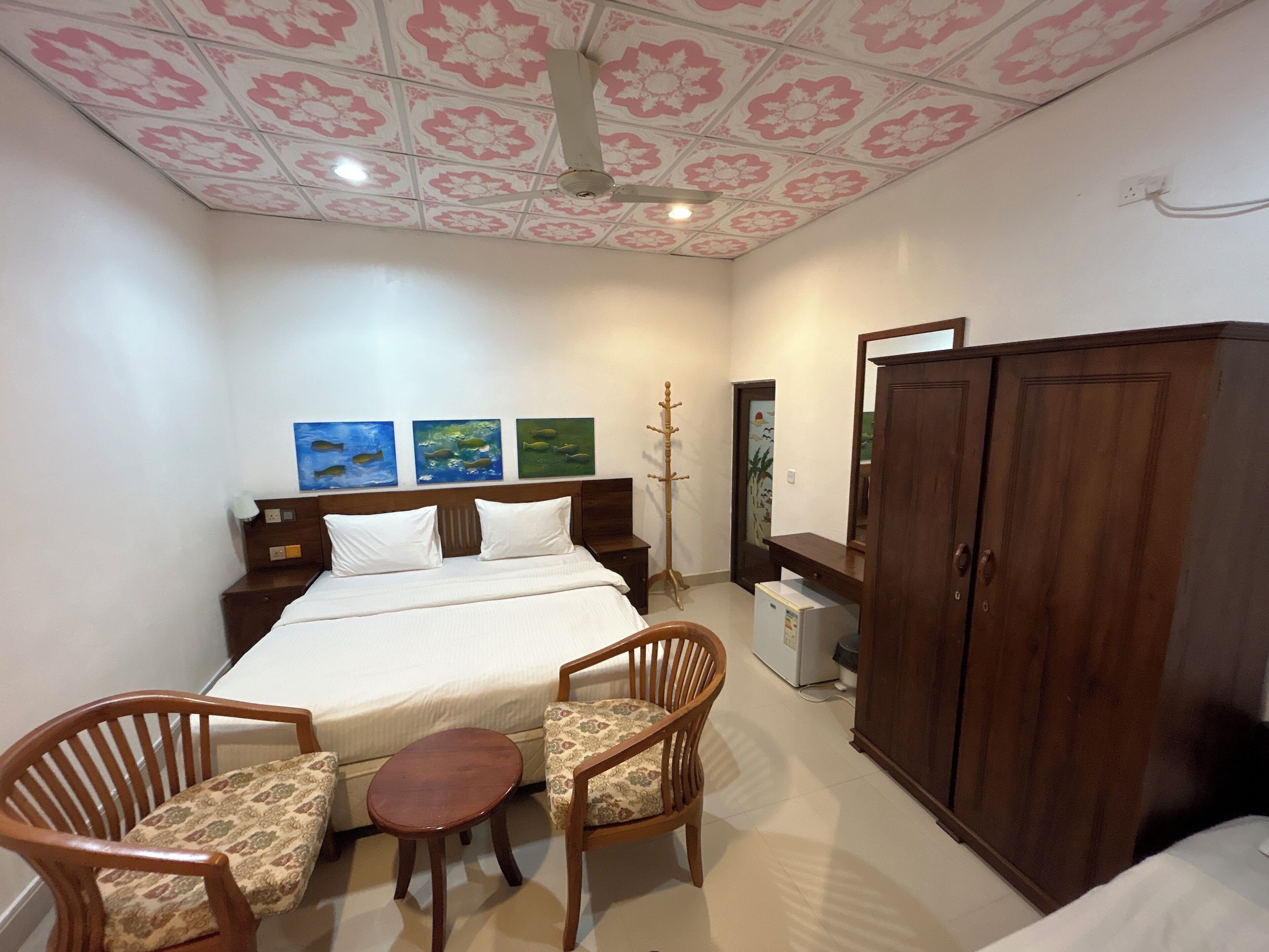 Quarto de hotel com uma cama, duas cadeiras, uma mesinha, um guarda-roupa e três quadros na parede.