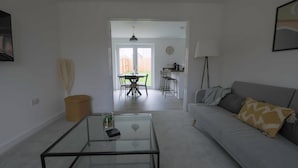 Uma sala de estar moderna com uma mesa de centro de vidro, um sofá cinzento, uma área de jantar com mesa e cadeiras, e uma cozinha com um abajur.