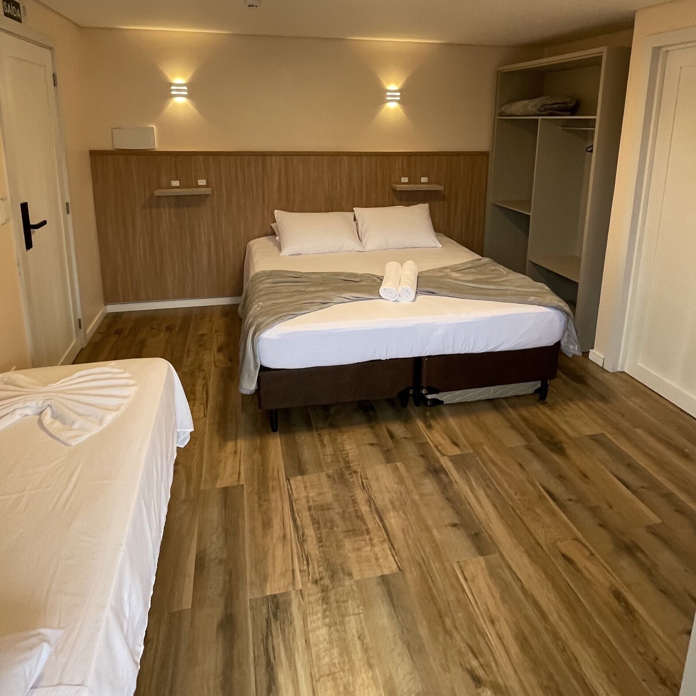 Quarto de hotel com cama, mesa de cabeceira, guarda-roupa e porta.