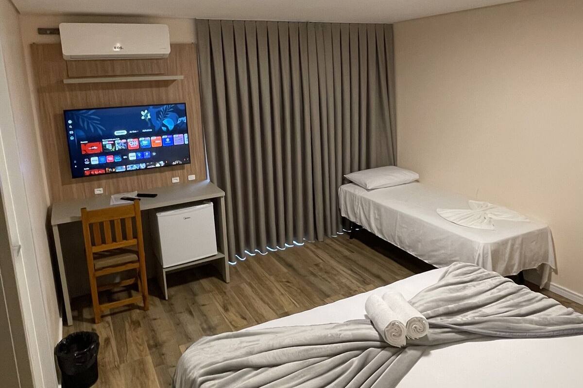 Quarto de hotel com duas camas, uma mesa, uma televisão e uma cortina.