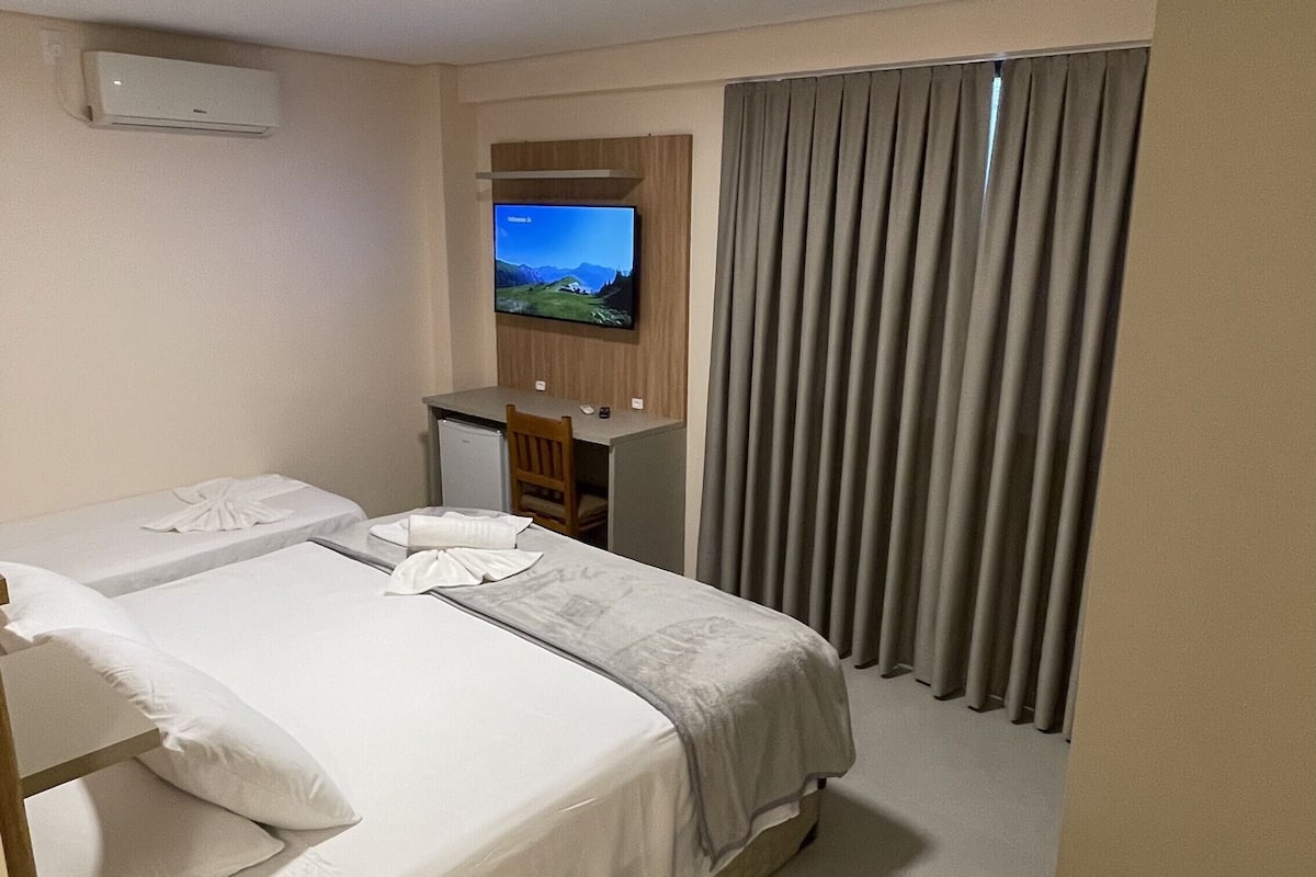 Quarto de hotel com duas camas, uma escrivaninha, uma televisão e janela com cortinas.