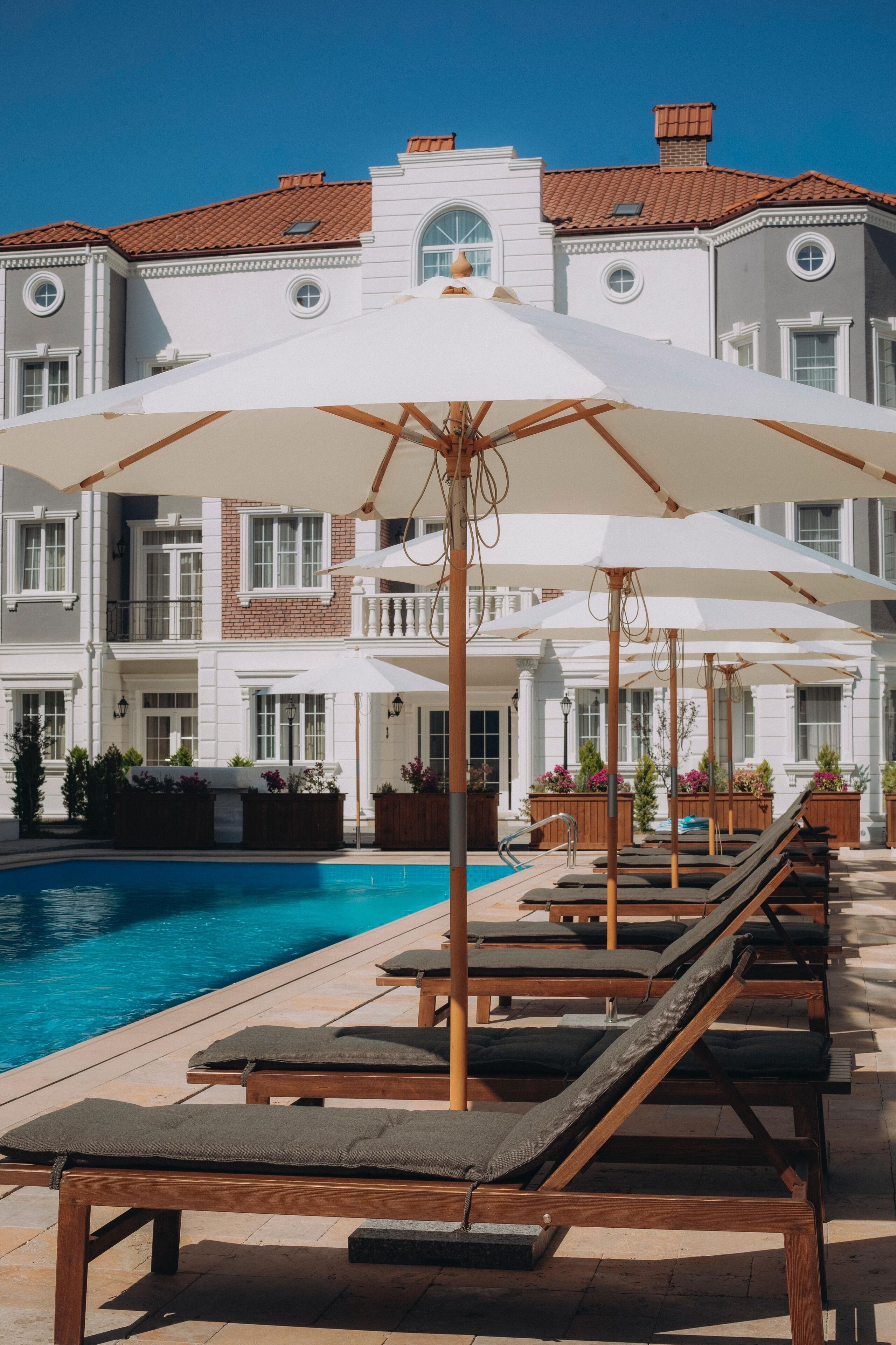 Uma área à beira da piscina com espreguiçadeiras e guarda-sóis, uma piscina e um edifício com telhado de telhas vermelhas e paredes brancas.