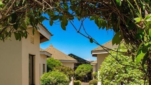 Uma passagem ladeada por sebes bem aparadas e uma fileira de edifícios brancos, sob um céu azul límpido.