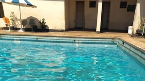 Piscina com água azul cristalina, área de estar com cadeiras e uma mesa, e um edifício com portas e janelas ao fundo.
