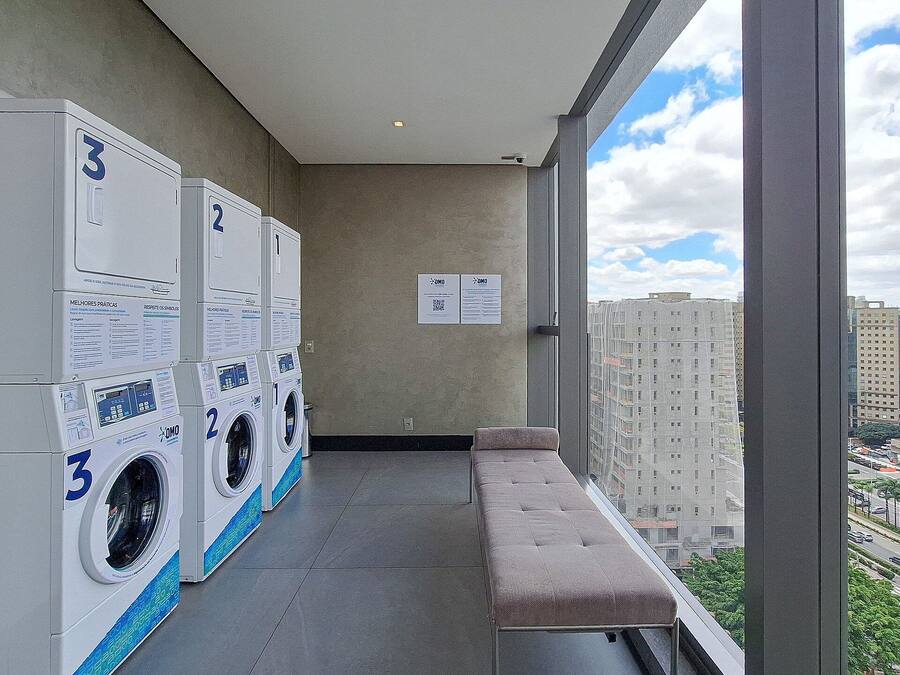 Uma lavanderia com três máquinas de lavar, um banco e vista para a cidade através de grandes janelas.