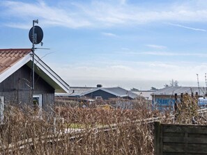 Paisagem rural com um edifício de madeira, antena parabólica, vegetação seca e outros edifícios ao fundo.