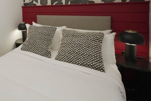 Cama com roupa de cama branca e duas almofadas estampadas, cabeceira vermelha e duas mesas de cabeceira com abajures.