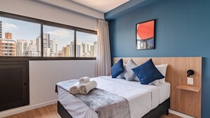 Quarto de hotel com uma janela grande, cama com roupa de cama azul e branca, cabeceira de madeira e vista para a cidade.