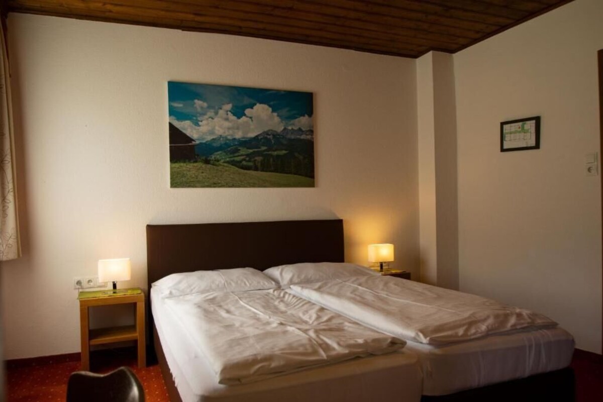 Quarto de hotel com cama, duas luminárias de cabeceira, um quadro de paisagem montanhosa e uma pequena mesa.